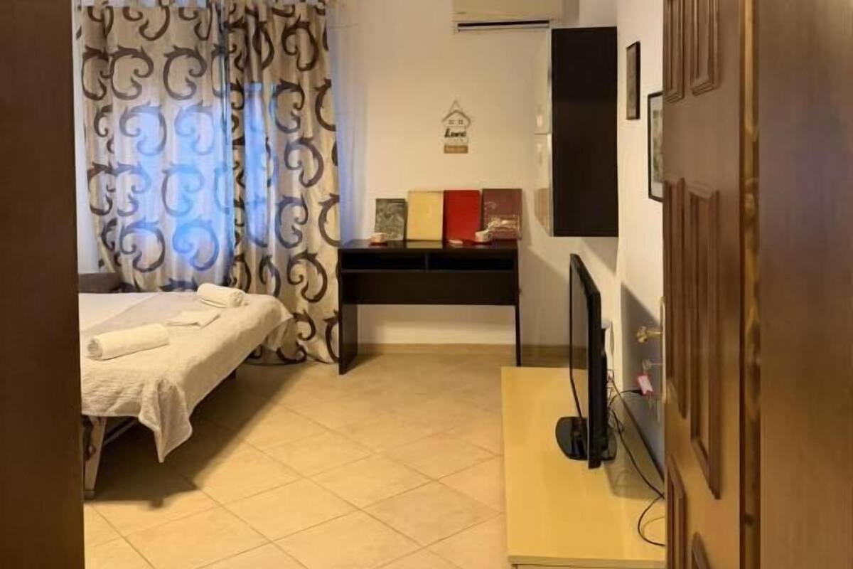 Quarto com cama, escrivaninha, televisão e ar condicionado.