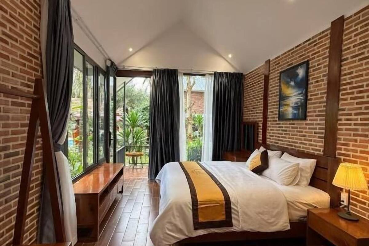 Quarto de hotel com cama, mesa de cabeceira, luminária e vista para o verde através de grandes janelas.