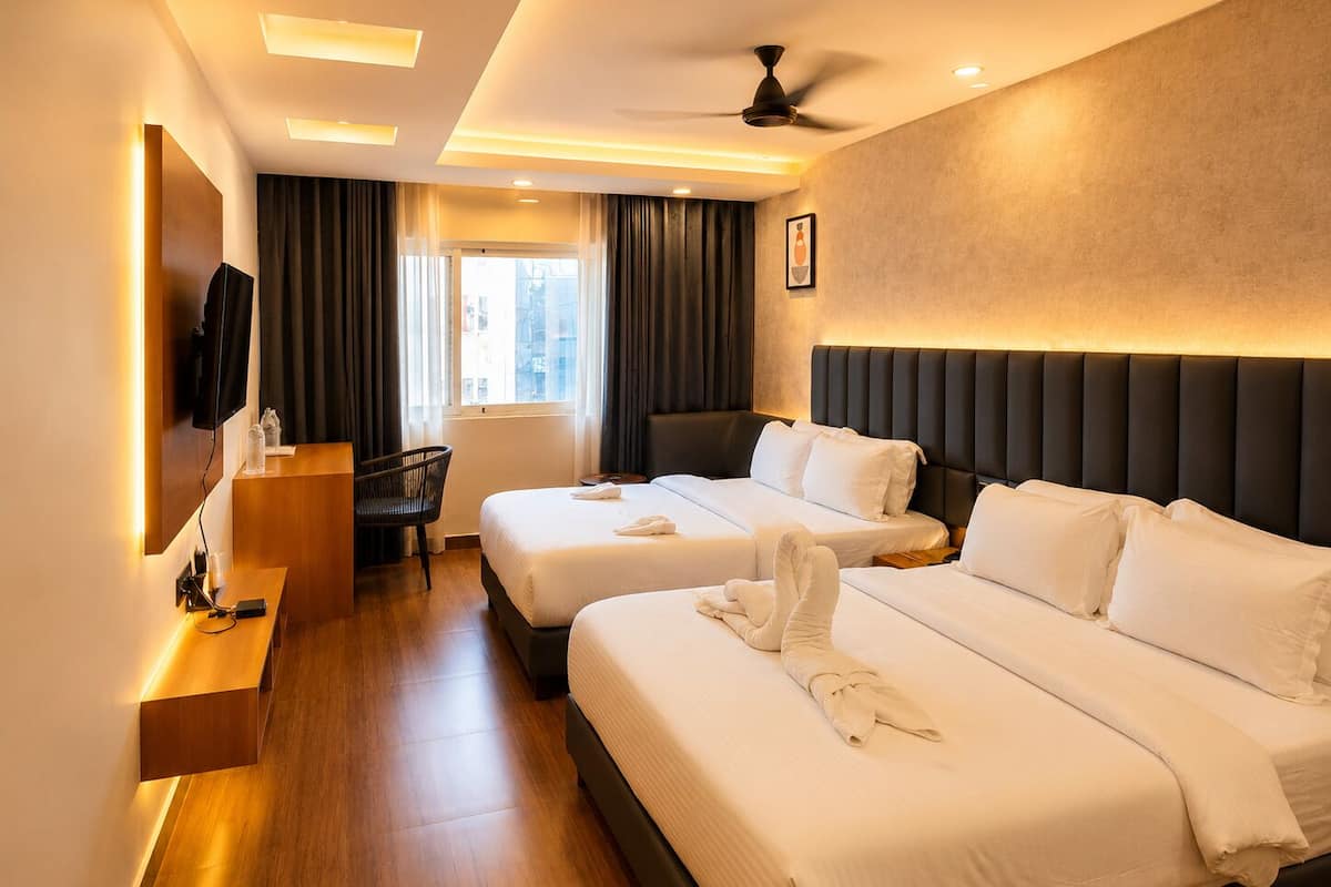 Quarto de hotel com duas camas, uma escrivaninha, uma cadeira, uma televisão e um ventilador de teto.