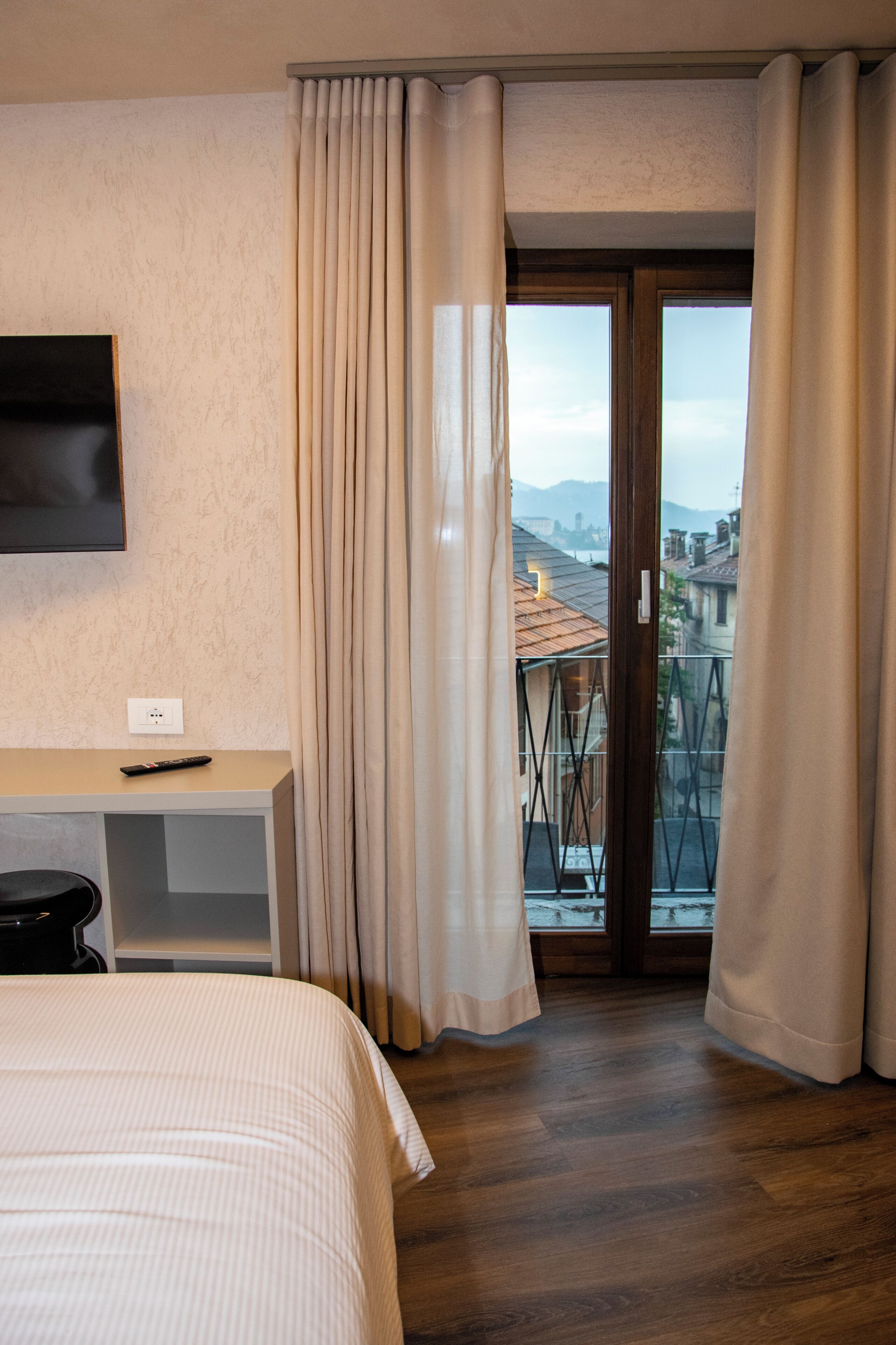 Quarto de hotel com cama, escrivaninha, televisão e varanda com cortinas.