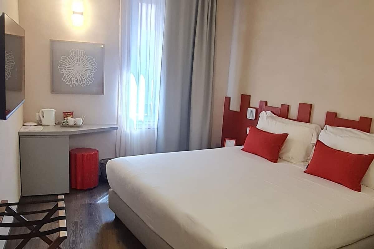 Quarto de hotel com uma cama grande, uma escrivaninha, uma cadeira e um banquinho vermelho.