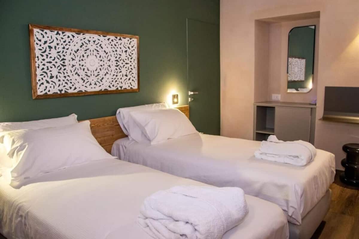 Quarto de hotel com duas camas, cabeceira de madeira, painel decorativo na parede, espelho e uma pequena escrivaninha com luminária.