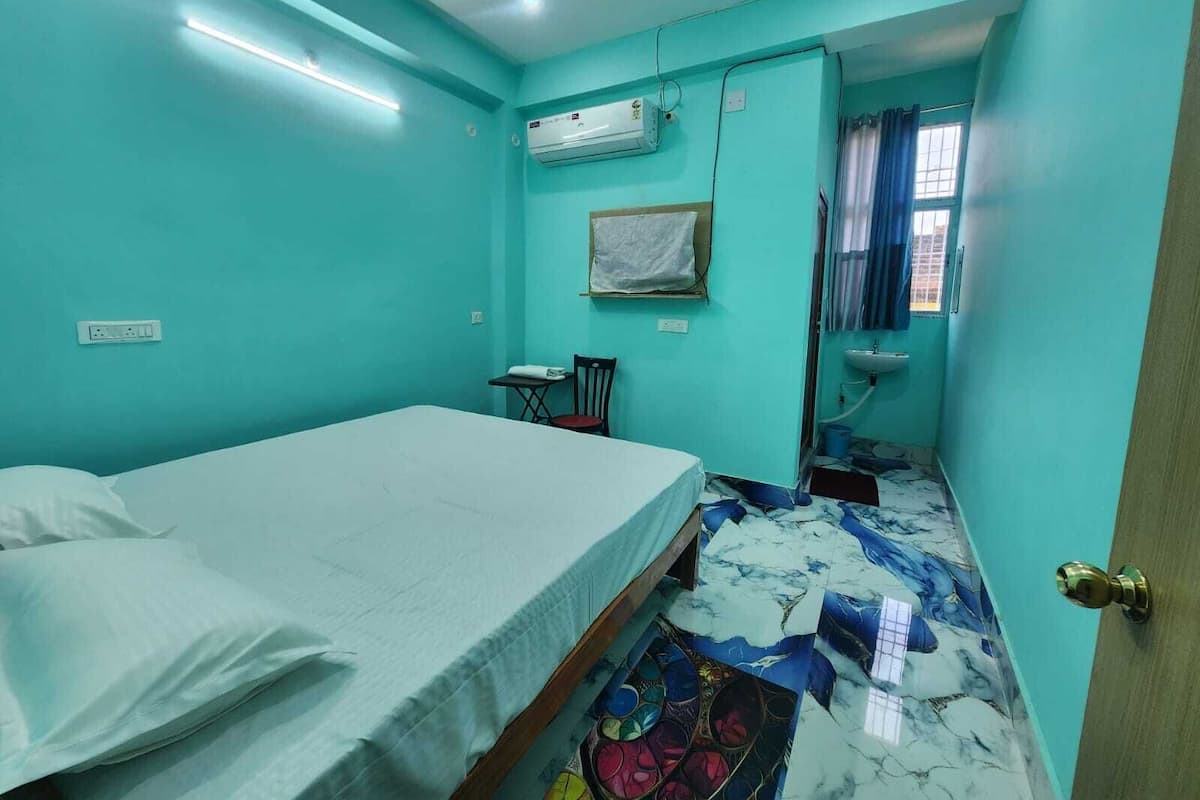 Quarto com paredes verde-água, cama com roupa de cama branca, ar condicionado, mesa pequena com uma cadeira, pia e chão estampado.