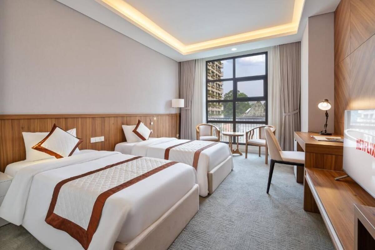 Quarto de hotel com duas camas, uma escrivaninha, uma cadeira, televisão e uma janela grande com vista.