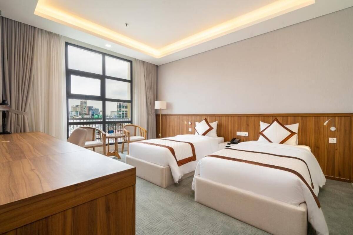 Quarto de hotel com duas camas, uma escrivaninha, uma cadeira e vista da cidade através de amplas janelas.