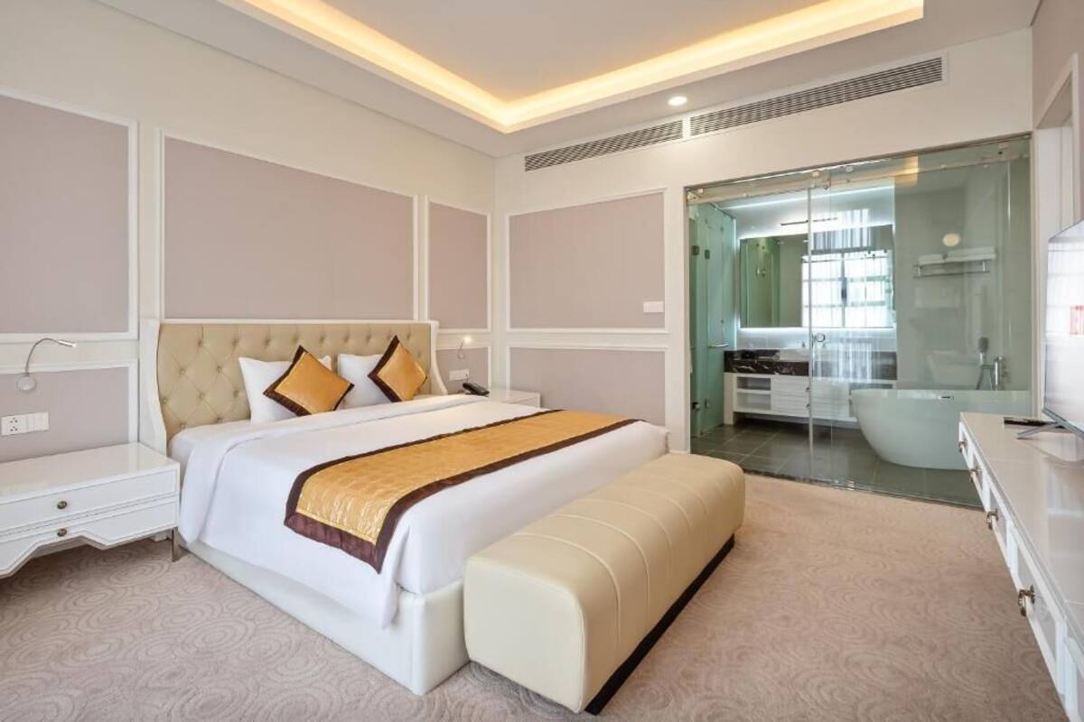 Quarto de hotel moderno com uma cama grande, um banco, uma toucador e uma casa de banho envidraçada.