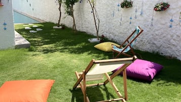 Um quintal com uma cadeira de madeira no deck, uma piscina pequena e uma parede branca com detalhes decorativos.