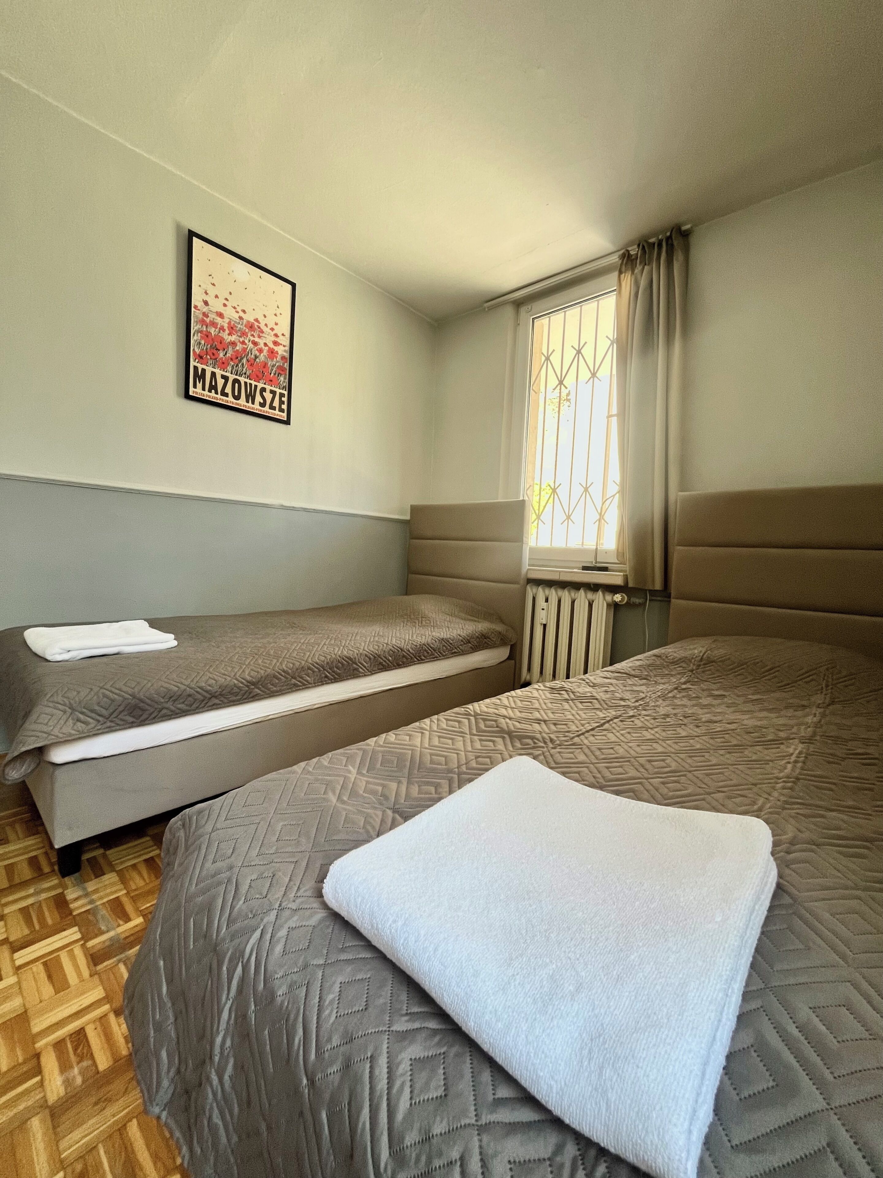 Quarto de hotel com duas camas, um radiador, uma janela com cortinas e um quadro na parede.