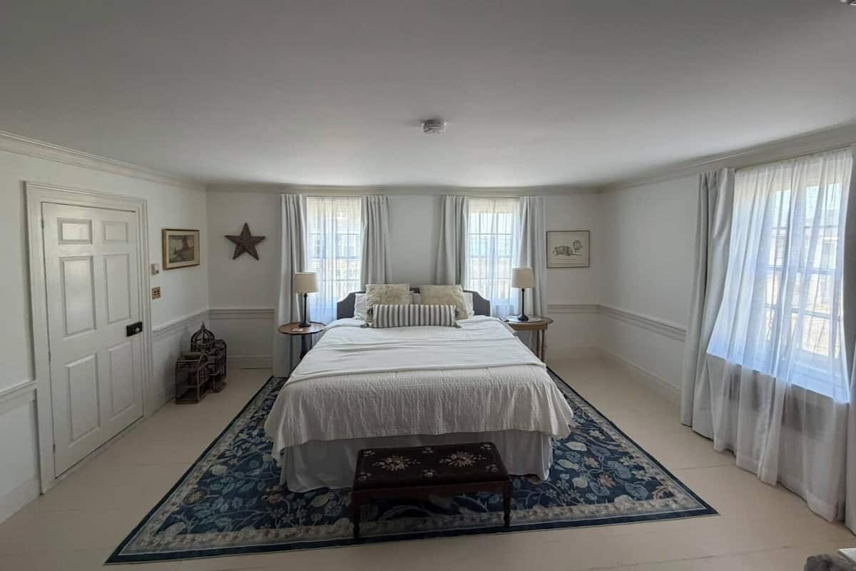 Quarto com cama grande, duas mesas de cabeceira, radiador e janela com cortinas.