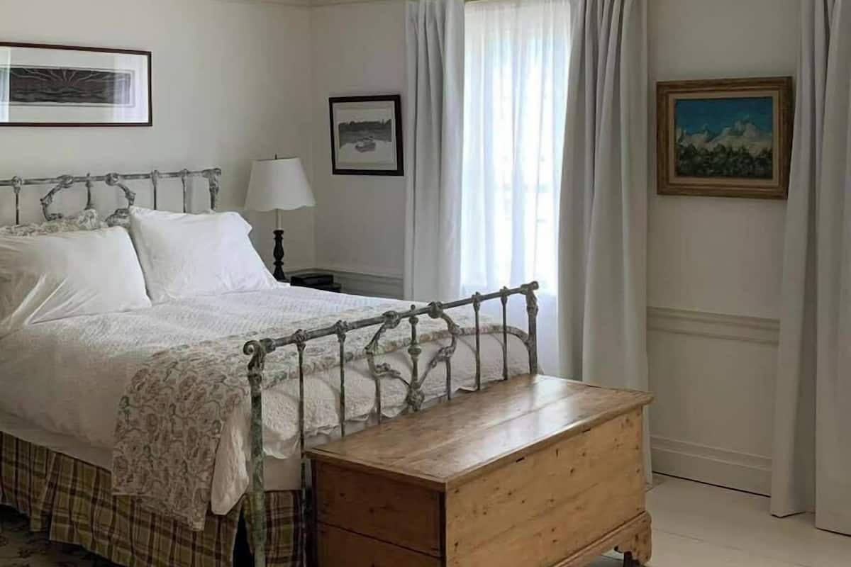 Um quarto com uma cama, um baú de madeira, uma mesa de cabeceira, um abajur e duas fotos emolduradas na parede.