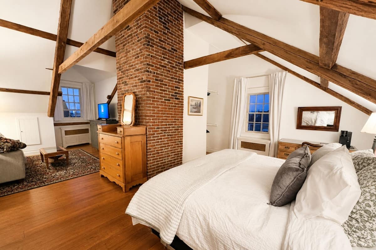 Quarto com lareira de tijolos, vigas de madeira, cama com roupa de cama branca, cômoda e escrivaninha com computador.