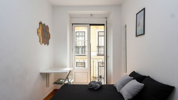 Quarto com cama, almofadas, uma mesinha e janela com vista para varanda.