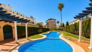 Uma piscina com design curvo, rodeada por um deck ladrilhado, palmeiras e um edifício com uma placa que diz 'CASTELL'.