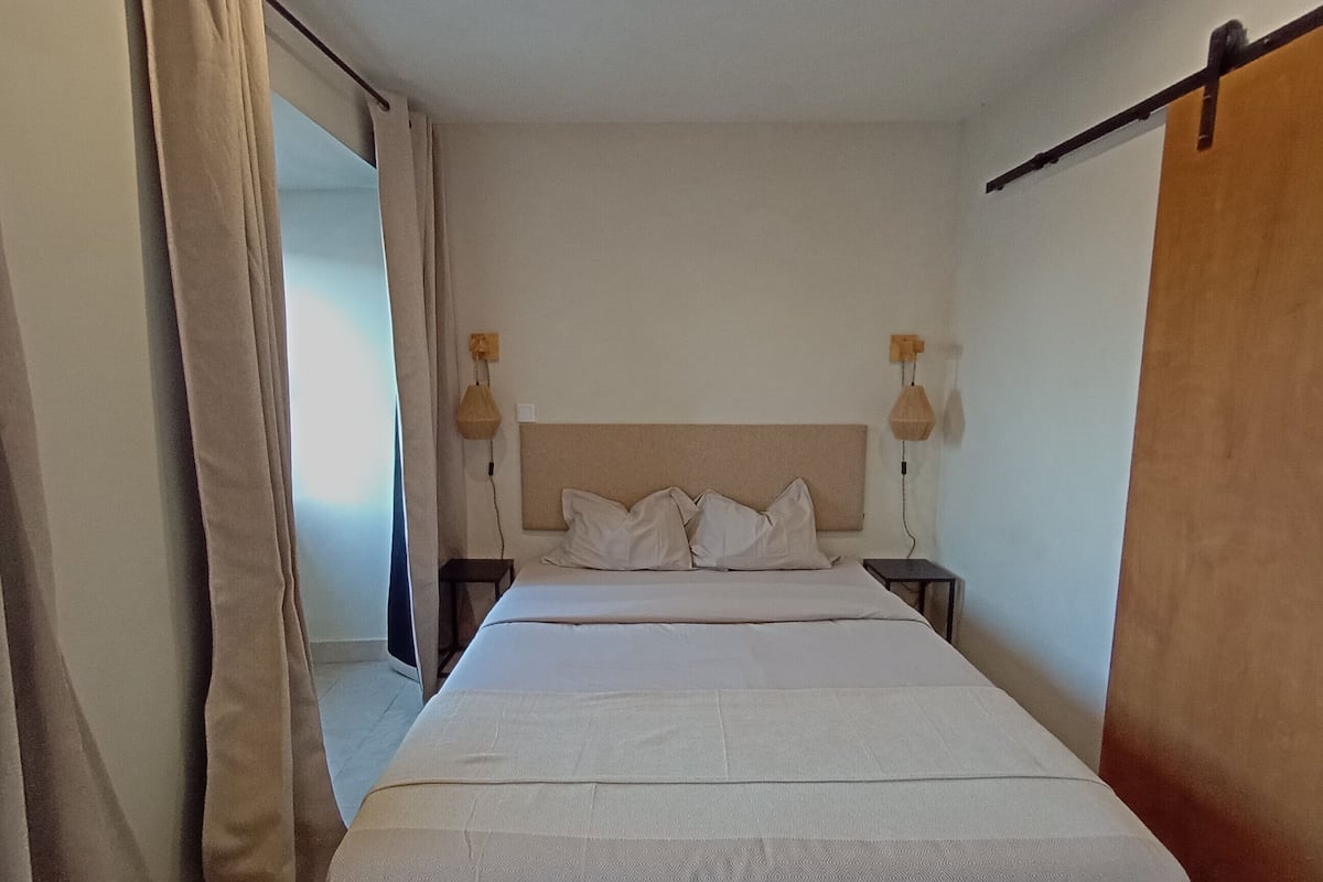 Quarto com cama, duas mesas de cabeceira, encosto e janela com cortinas.