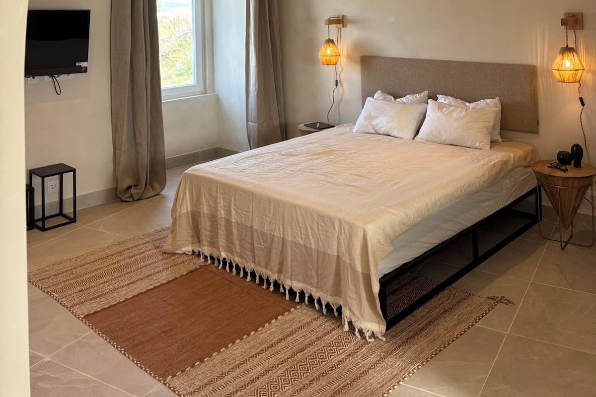 Quarto com cama, duas luminárias de cabeceira, televisão e janela com cortinas.