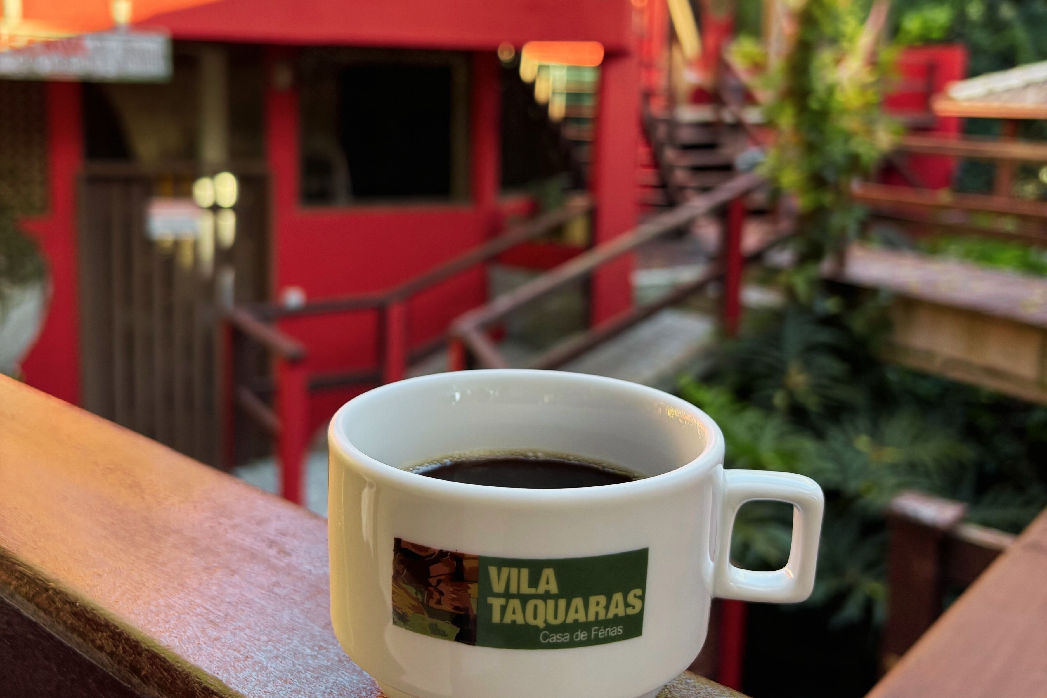 Uma caneca branca com a inscrição 'Vila Taquaras Casa de Fênas', colocada numa varanda com um edifício vermelho ao fundo.