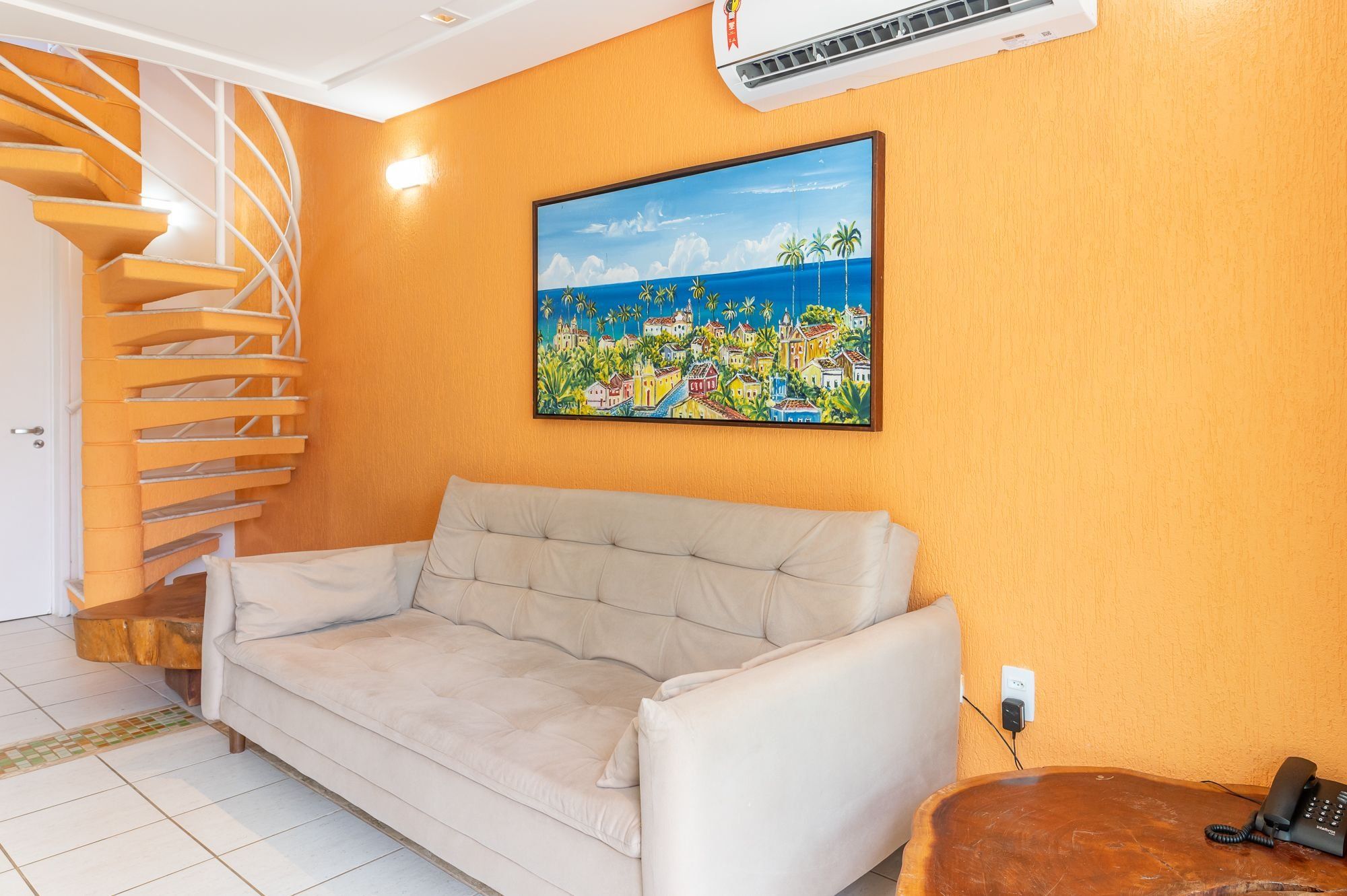 Um quarto com um sofá bege, uma pintura de uma cidade costeira, uma escada em espiral e um aparelho de ar condicionado.