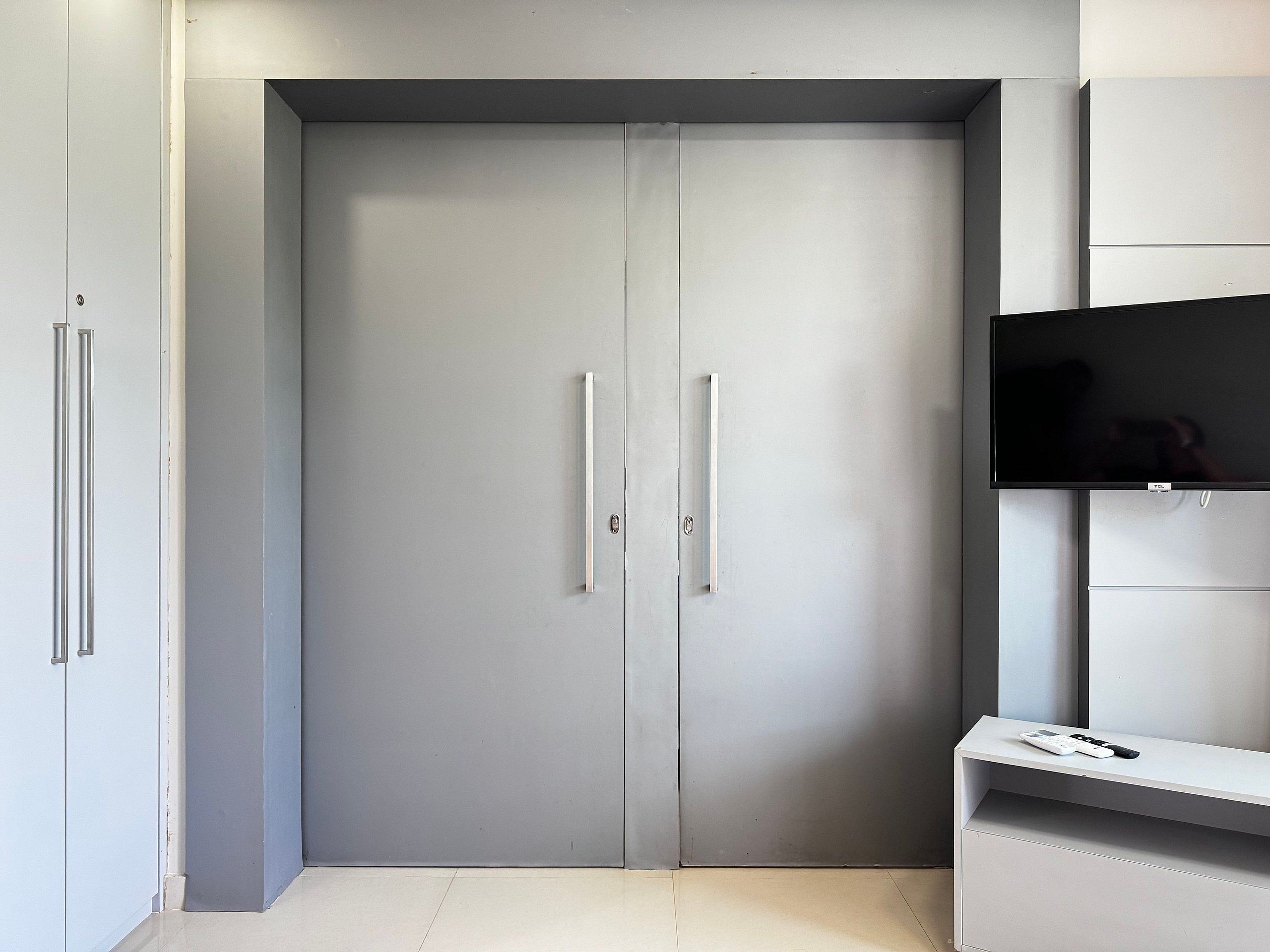 Quarto moderno com guarda-roupa cinzento, televisão de tela plana na parede e uma pequena estante branca com controle remoto.