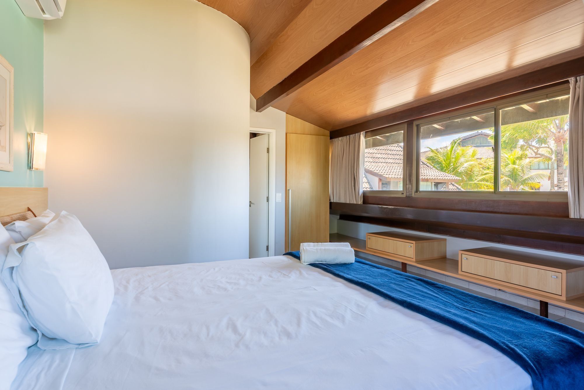 Quarto de hotel com cama, janela e vista para palmeiras, e teto de madeira.