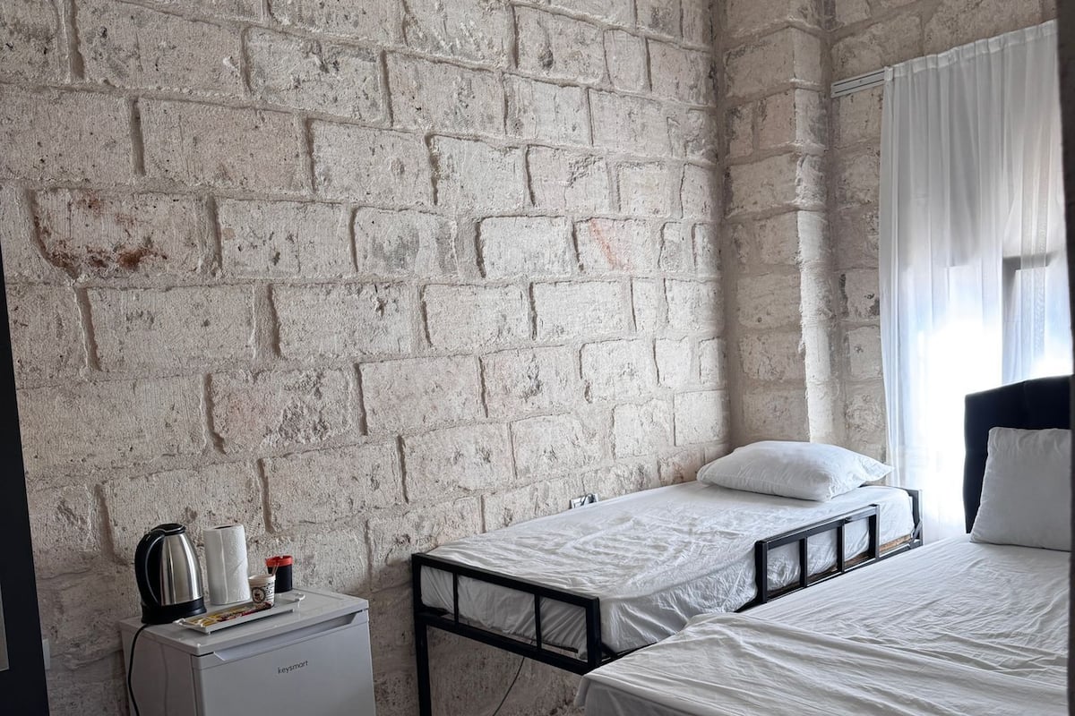 Quarto com parede de tijolos, cama, um pequeno frigorífico e janela com cortinas.