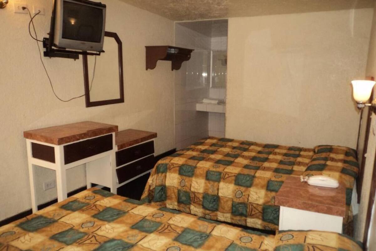 Quarto com cama, escrivaninha, televisão presa na parede e uma pequena estante.