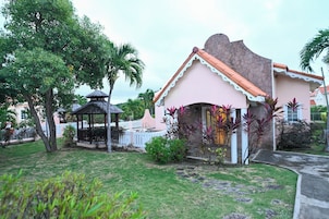 Um edifício rosa com fachada de pedra e telhado de telhas, rodeado por uma cerca branca de piquete, palmeiras e um coreto.