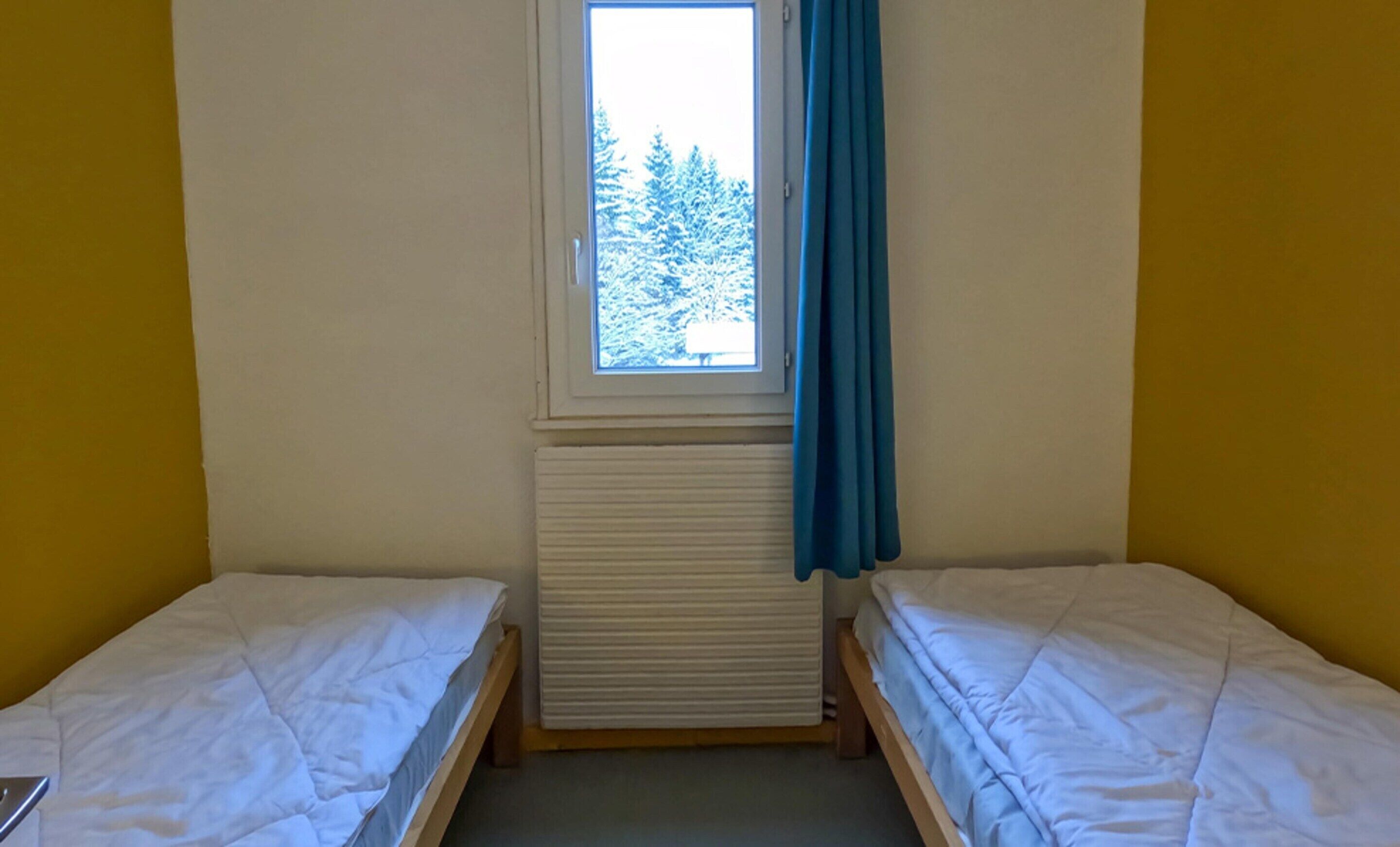 Quarto pequeno com duas camas, uma janela com vista para árvores e um radiador.