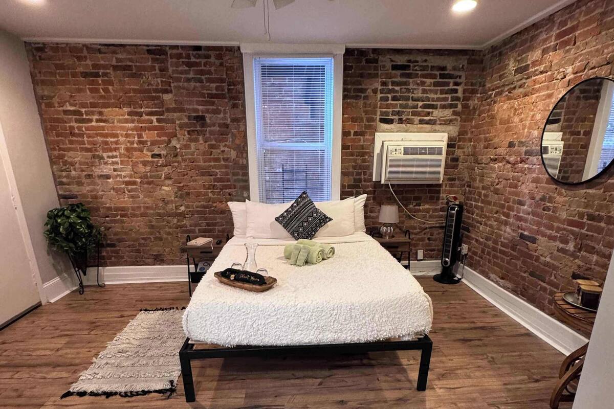 Um quarto com parede de tijolos, cama com roupa de cama branca, ventilador de teto, janela com persianas e piso de madeira.