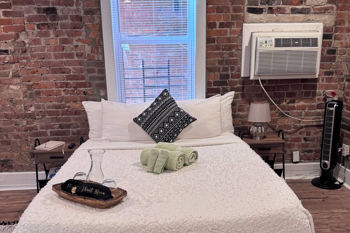 Cama com cobertura branca e fofa, travesseiro estampado e toalhas enroladas. Pequeno tabuleiro de madeira com planta e garrafa. Janela com persianas visíveis, e ar condicionado fixado na parede de tijolos.