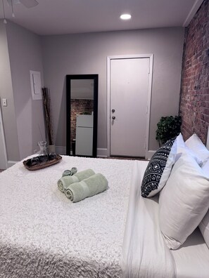 Quarto com cama, travesseiros, mesa de cabeceira e um copo sobre ela, e uma porta.