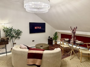 Uma sala de estar moderna com sofá, mesa de jantar, lustre e TV exibindo 'NETFLIX'.