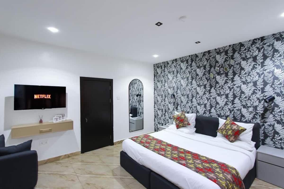 Quarto de hotel moderno com uma cama grande, uma televisão fixada na parede, uma mesinha de cabeceira e um papel de parede estampado.