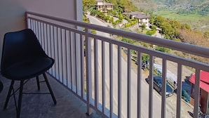 Uma varanda com uma cadeira, vista de um vale com casas e montanhas, e céu claro.