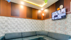 Quarto nos fundos com sofá modular, paredes de madeira, papel de parede estampado e uma TV fixada exibindo imagens de segurança.