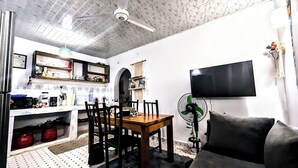 Uma sala de jantar com mesa e cadeiras de madeira, uma cozinha com prateleiras e eletrodomésticos, um ventilador e uma TV fixada na parede.
