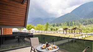 Varanda com mesa de vidro posta para o café da manhã, com vista para uma paisagem montanhosa com iurtes e construções de madeira.