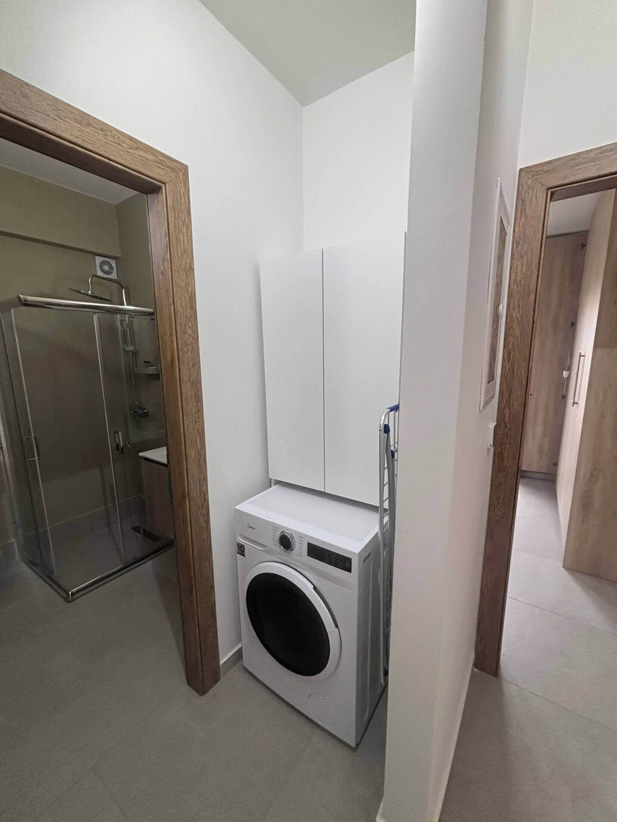 Uma lavanderia compacta com máquina de lavar, box de vidro para chuveiro e porta de madeira.