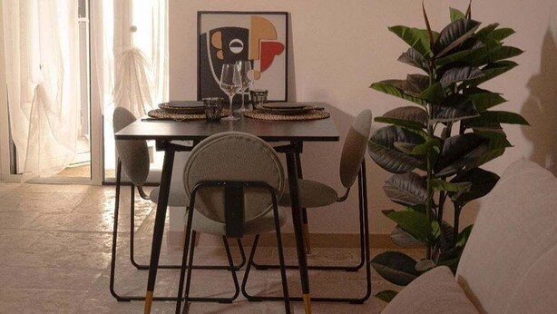 Uma área de refeições com uma mesa posta para dois, cadeiras, uma planta e uma obra de arte emoldurada na parede.