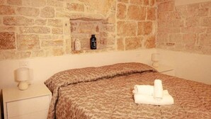 Quarto com parede de pedra, cama com colcha estampada, mesa de cabeceira com abajur e toalha sobre a cama.