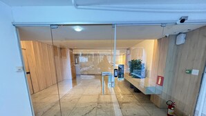 Uma área de recepção com porta de vidro, um extintor de incêndio, uma placa com a inscrição 'EMPURENCE' e um tapete de entrada com a inscrição 'RESIDENCIAL SAN CARLO'.