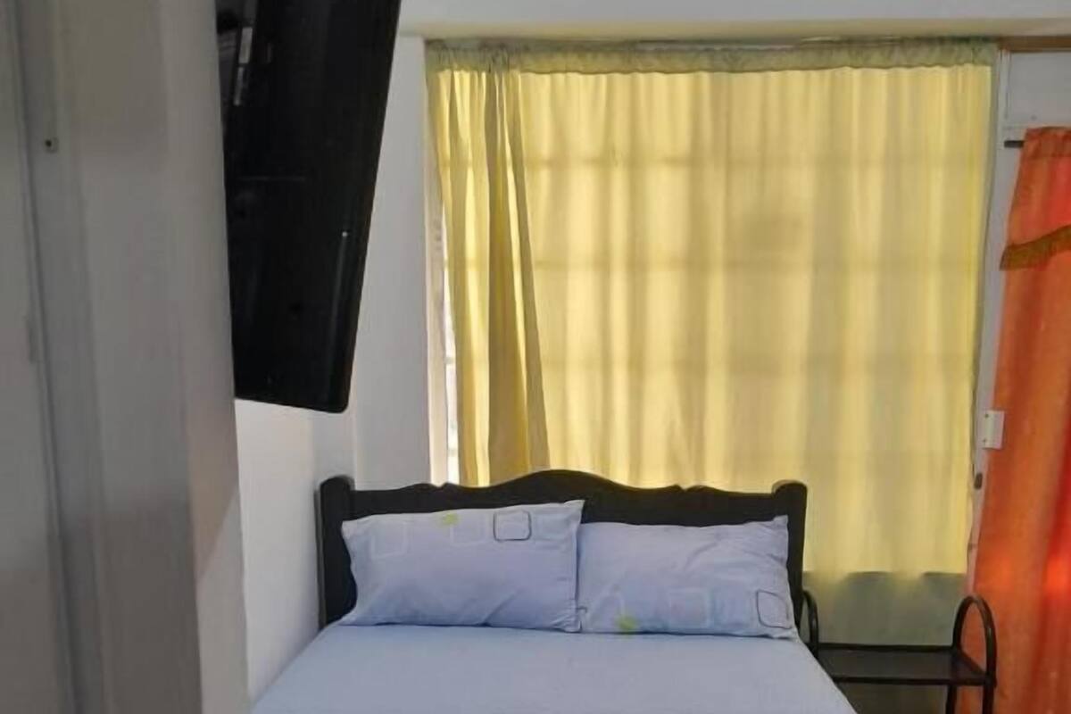 Cama com colcha azul, televisão fixada na parede e janela com cortinas amarelas.