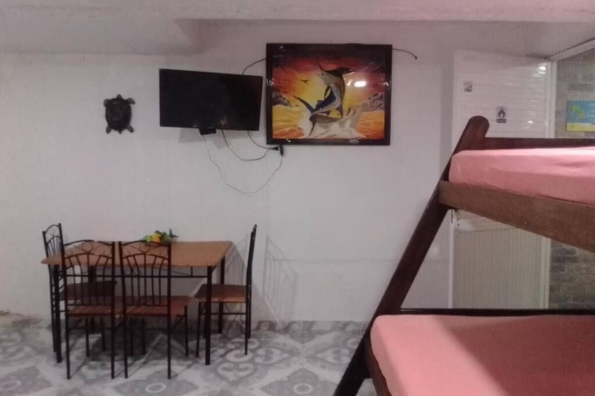 Quarto com cama de solteiro, mesa de jantar com cadeiras, televisão presa à parede e um quadro emoldurado.