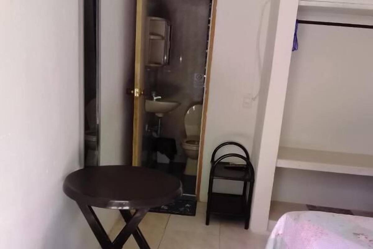 Um quarto pequeno com uma cama, um banquinho, uma mesa e uma porta para um banheiro.