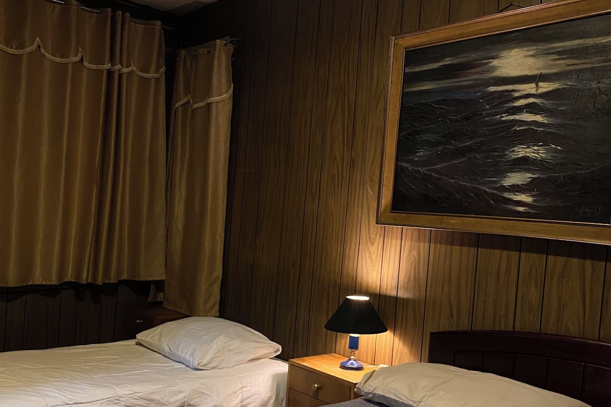 Quarto com duas camas, mesa de cabeceira com luminária, painéis de madeira e uma pintura na parede.