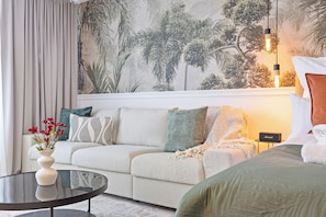 Quarto de hotel moderno com sofá, mesa de centro redonda de vidro, luminária fixada na parede e pintura com folhagem tropical.