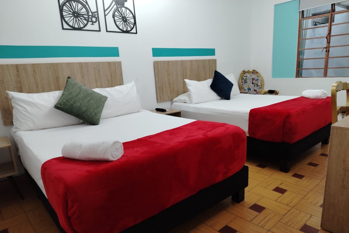 Quarto de hotel com duas camas, cada uma com colcha vermelha e almofadas brancas, cabeceira de madeira e janela com vista.