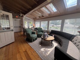 Um quarto iluminado pelo sol, com teto de madeira, um sofá, duas poltronas, uma mesa de centro e uma porta com a indicação 'EXIT'.
