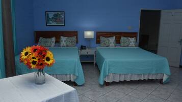Quarto de hotel com duas camas, uma mesa de cabeceira com abajur, uma mesa com um vaso de flores e uma porta.