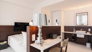 Quarto de hotel com cama, escrivaninha com luminária, cadeira, televisão, espelho, banheiro com pia e lixeira.