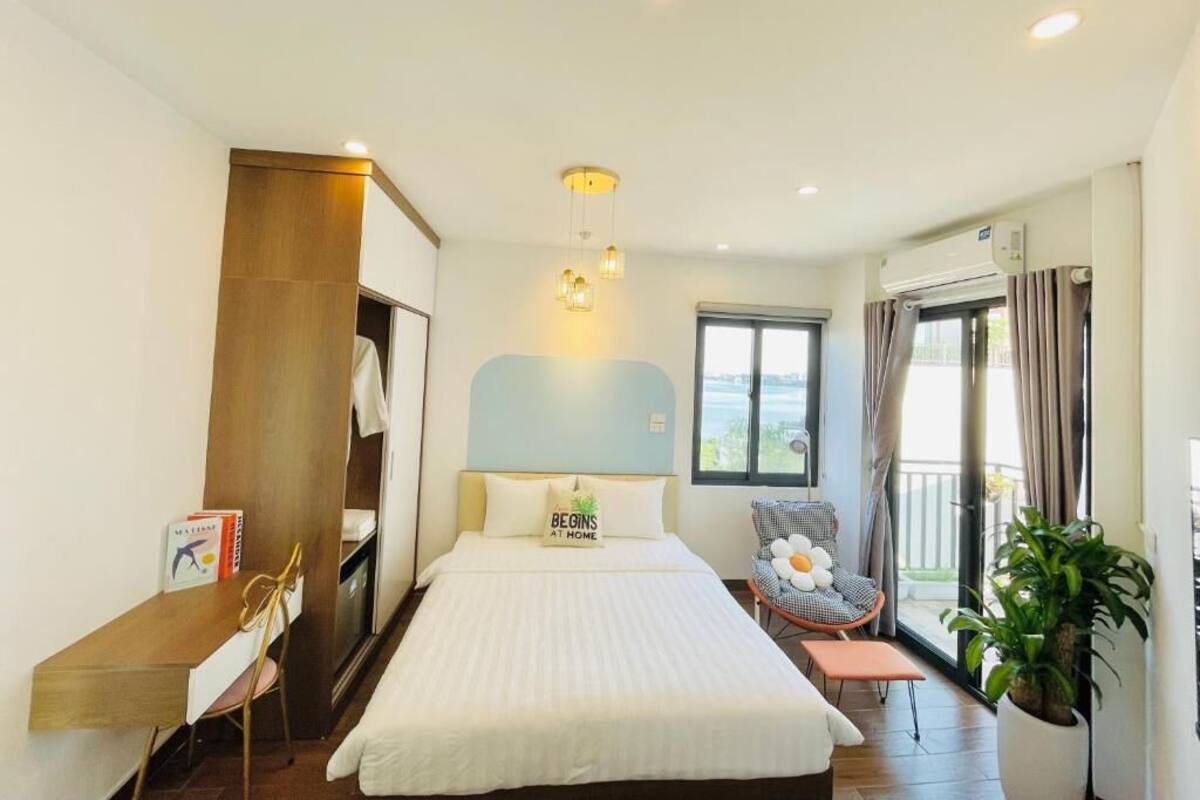 Quarto de hotel com cama, escrivaninha, cadeira, planta em vaso e vista para o exterior.